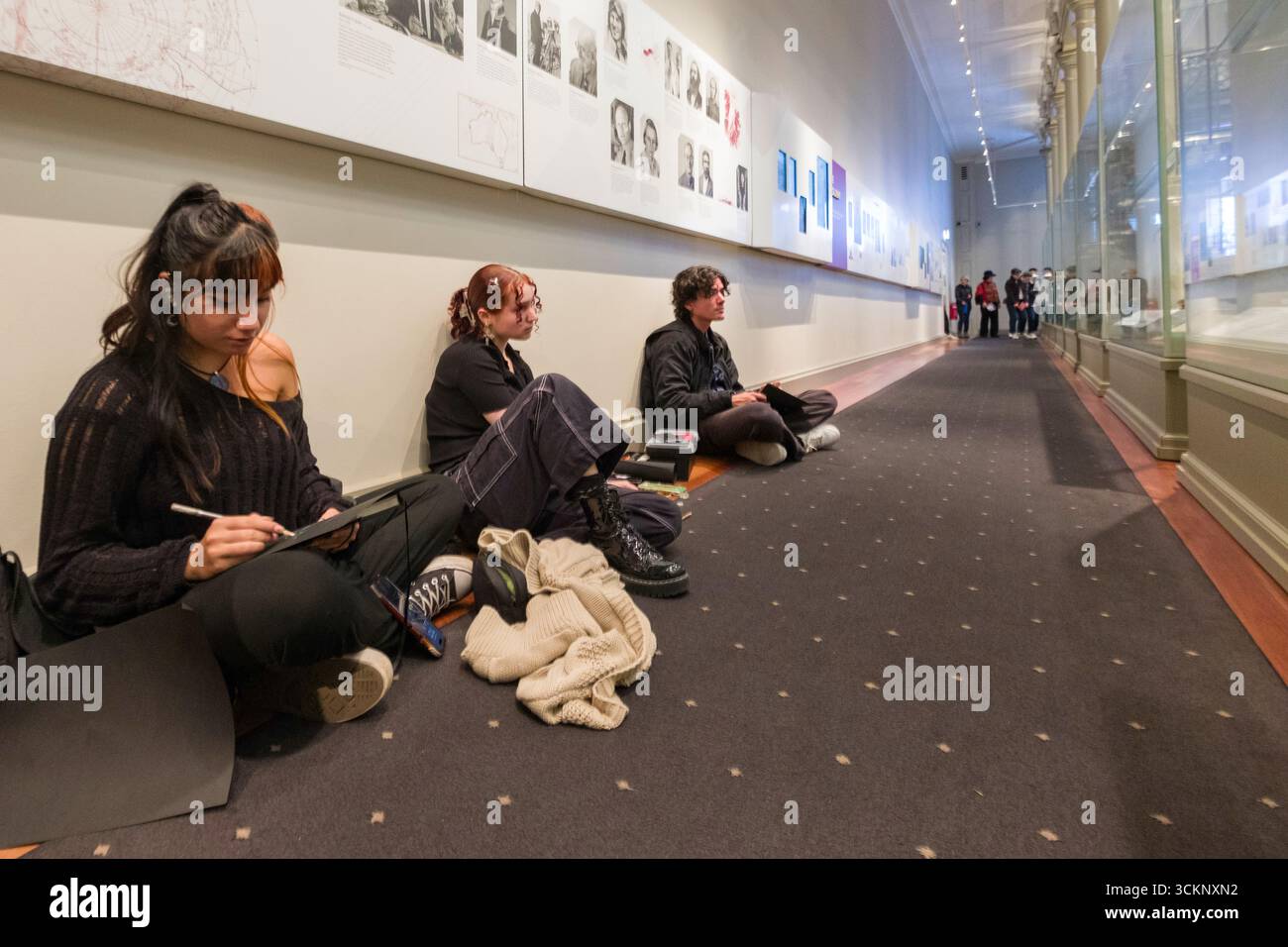 Several young individuals sit on the museum corridor floor, engaging in ...