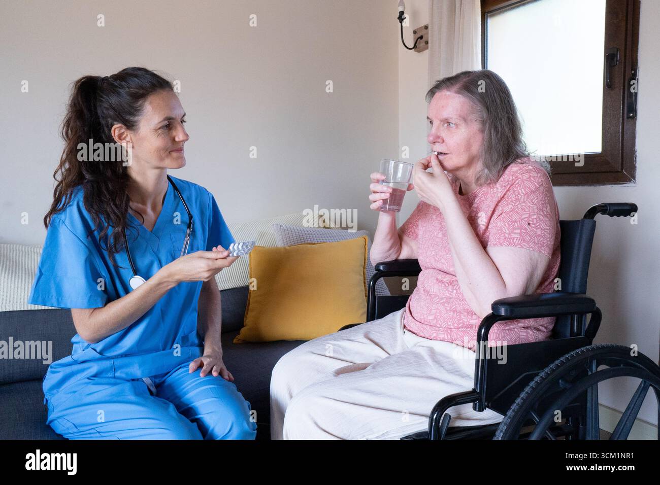 Nurse helps a senior woman in a wheelchair take her medication at home ...