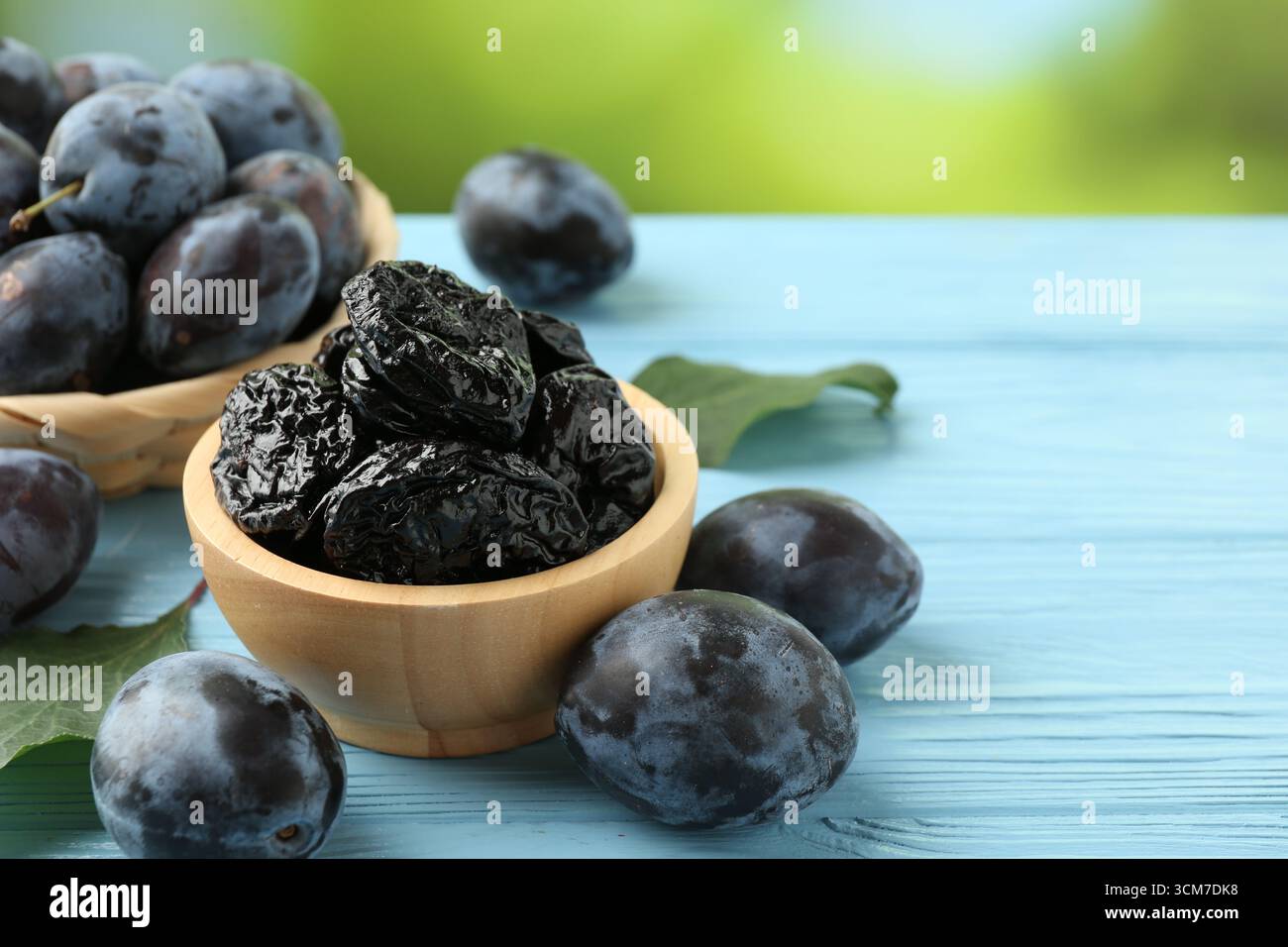 Dried prunes in bowl, leaves and fresh plums on light blue wooden table ...