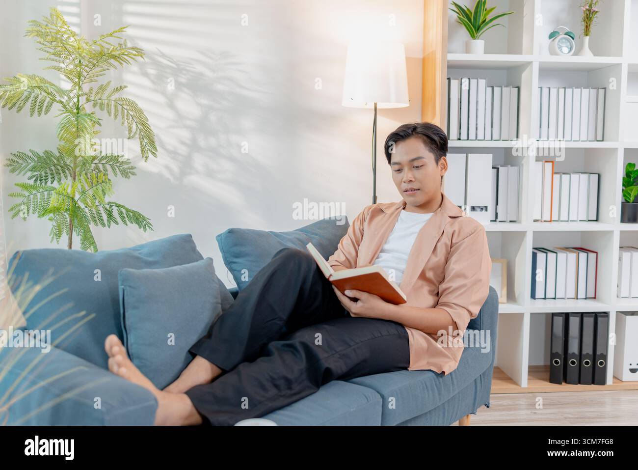 A young man sits comfortably on a sofa reading a book, appearing ...