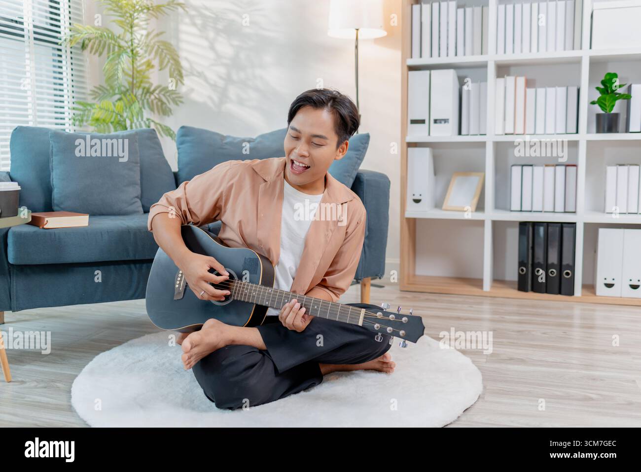 A young man sits cross-legged on the floor at home, joyfully playing an ...