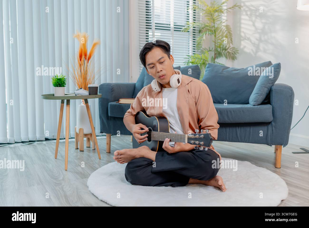 A young man sits cross-legged on the floor at home, joyfully playing an ...