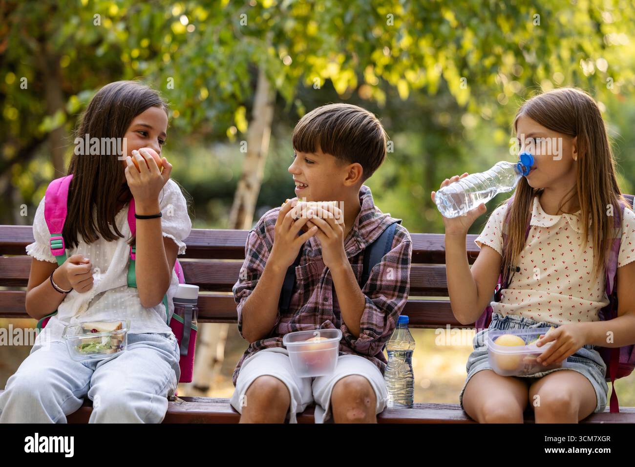 Three children are seated together i park, happily eating sandwiches ...