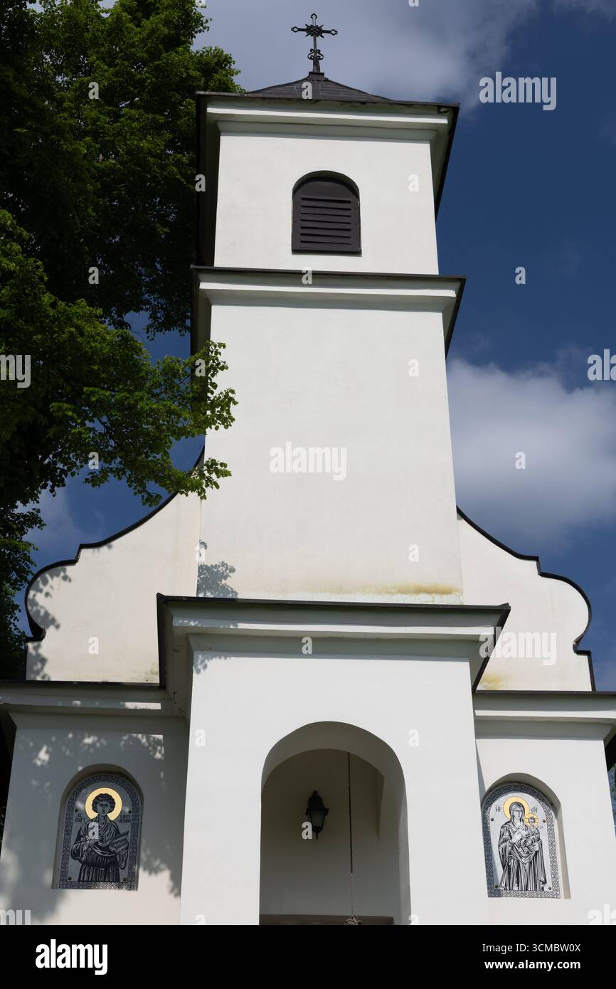 A white church tower with an overhead cross stands surrounded by lush ...