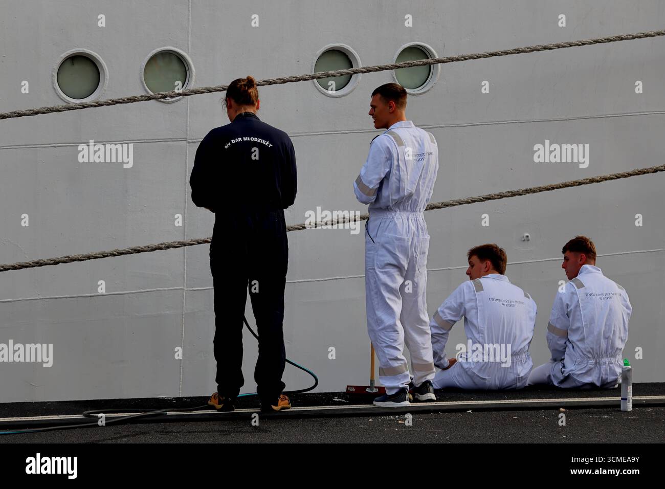 Crew in white coveralls sit and stand on the quay beside a moored tall ...