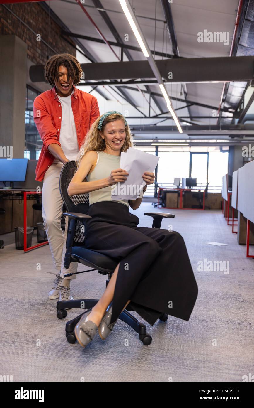 Diverse coworkers pushing rolling chair down aisle in open plan office ...
