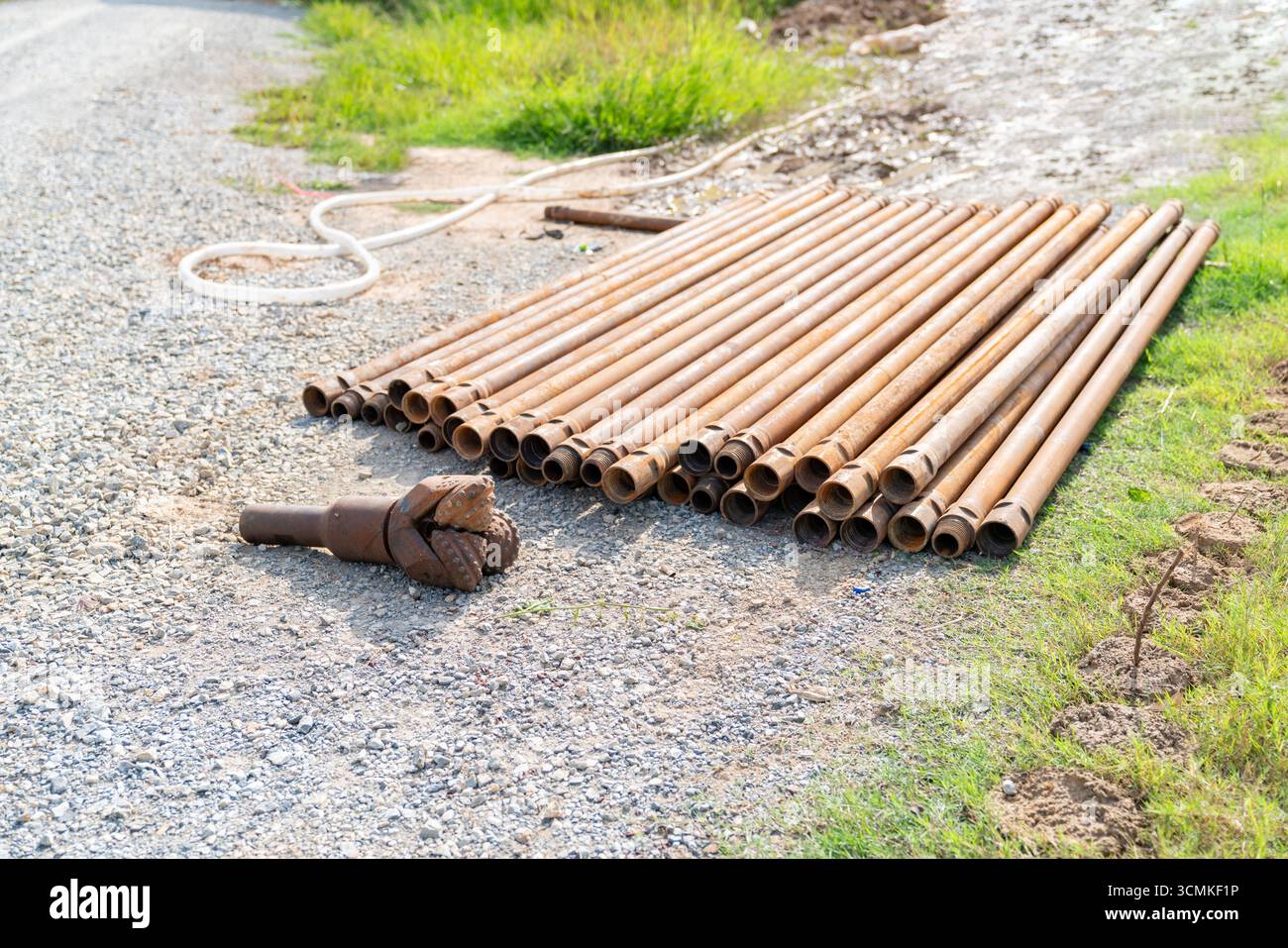 Rusty metal drill pipes and a tricone drill bit placed on gravel ground ...