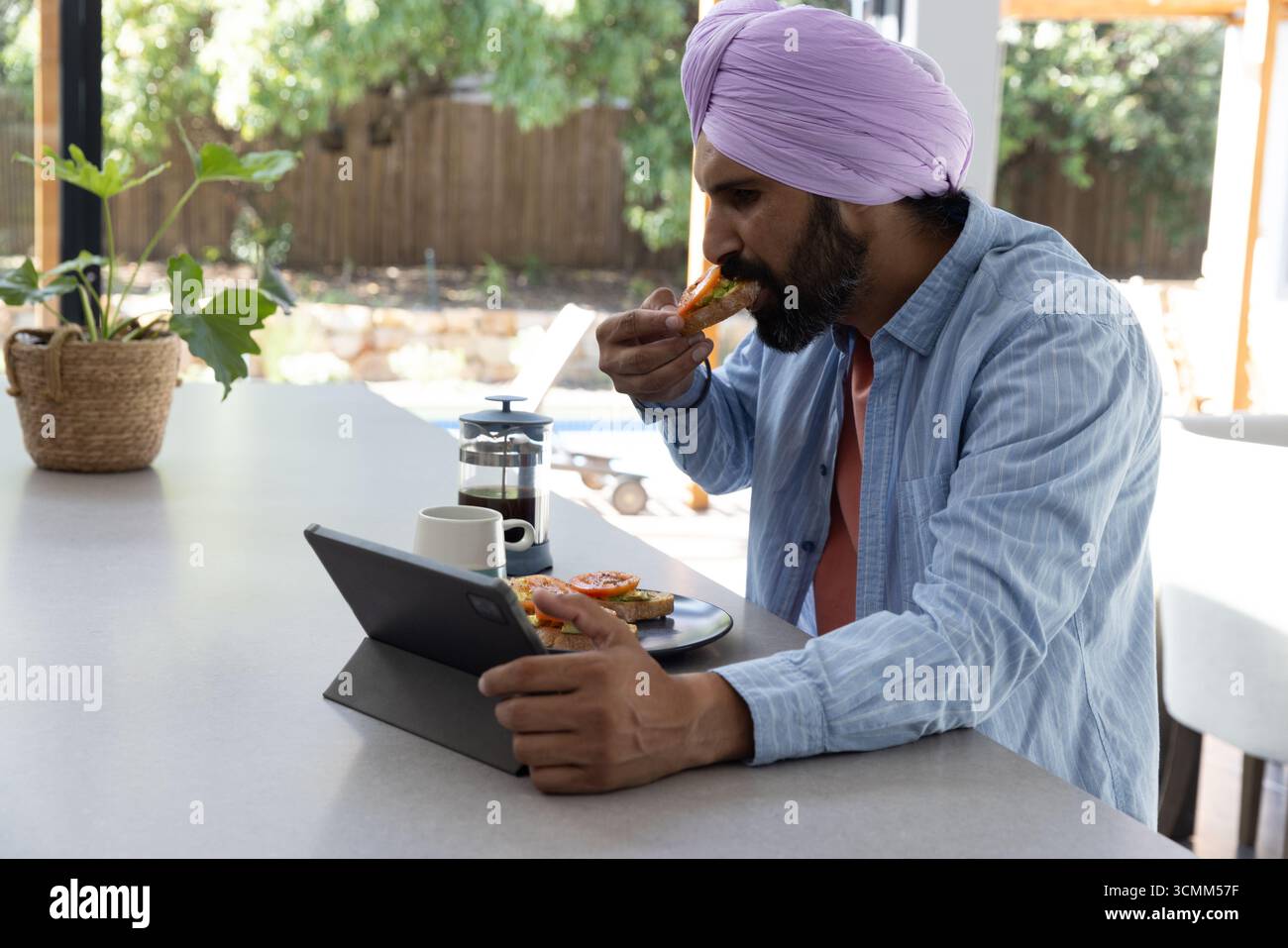 Indian man sitting at kitchen island using tablet and eating tomato ...