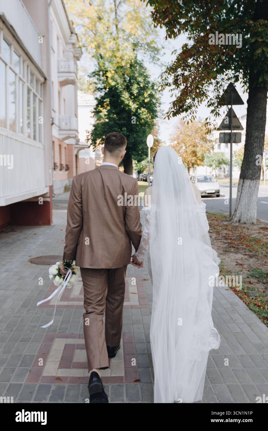 Bride and groom walk hand in hand along a city sidewalk, groom holding ...