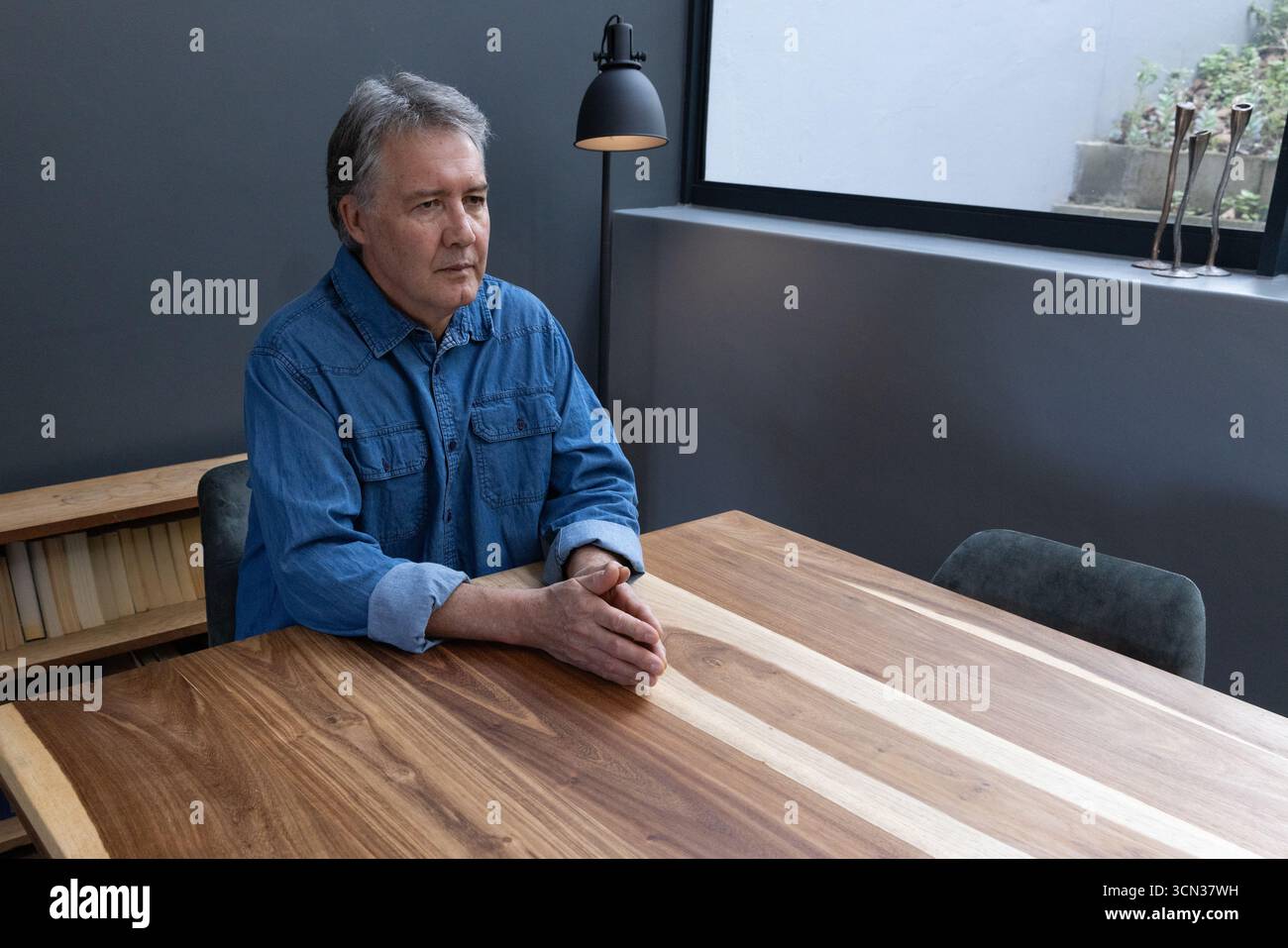 Senior man sitting at table with black lamp gazing at window in home ...