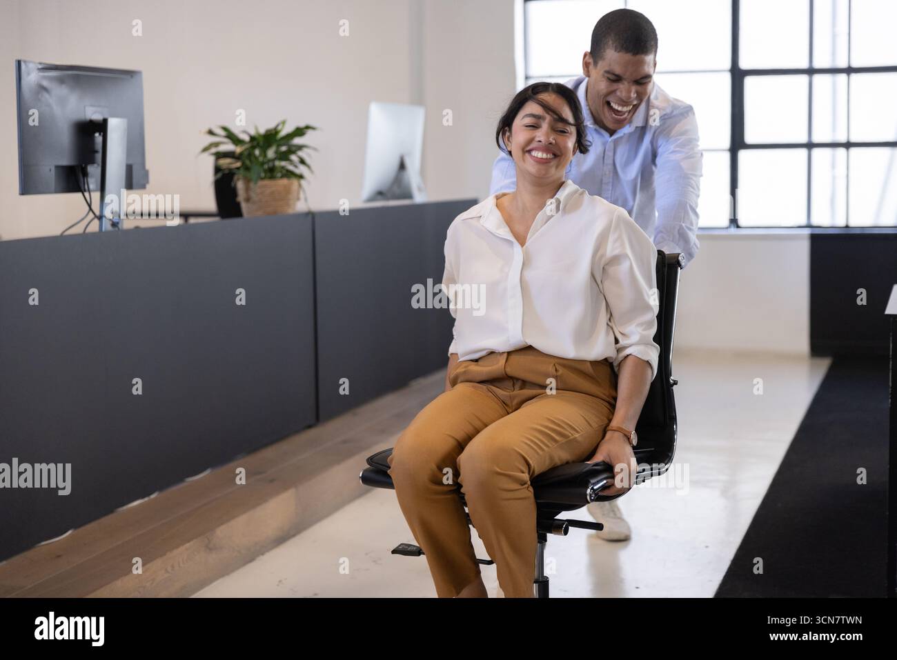 Diverse coworkers pushing swivel chair past desk partition, potted ...