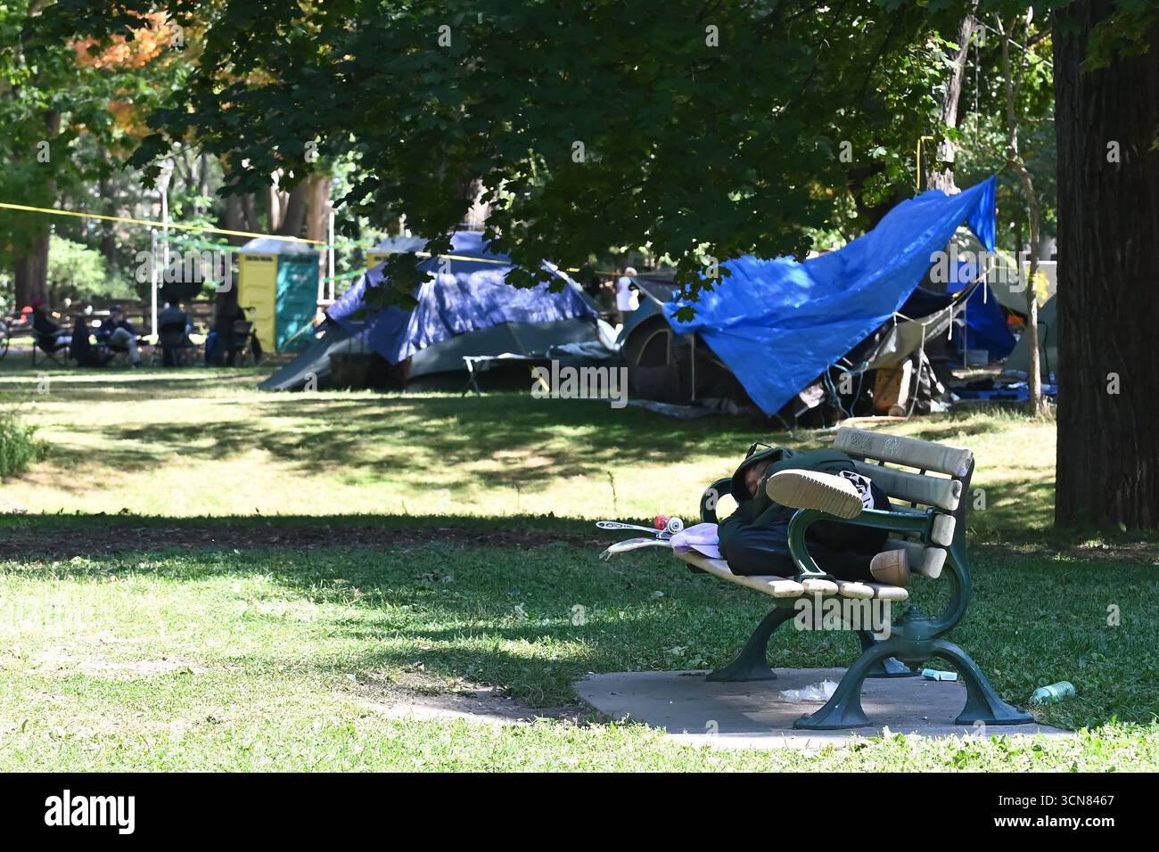 An unhoused person is seen resting on a bench near an encampment in ...