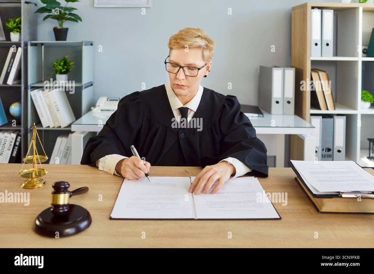 Female judge wearing robe writing document, scales of justice and gavel ...