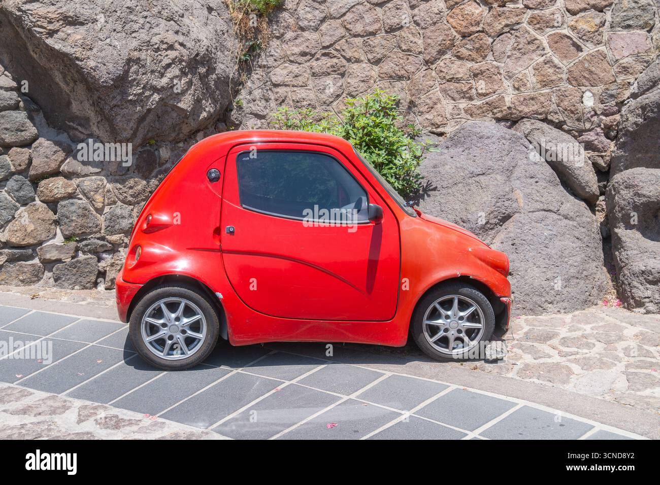Red microcar with visible damage is crashed into a stone wall, showing ...