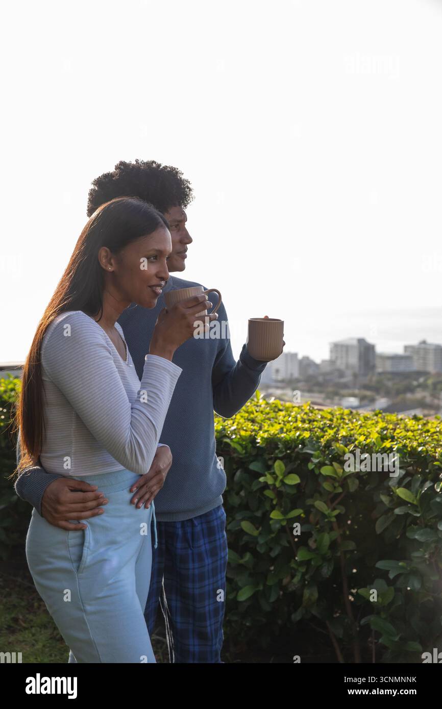 Indian couple standing on balcony with hedge holding mugs overlooking ...
