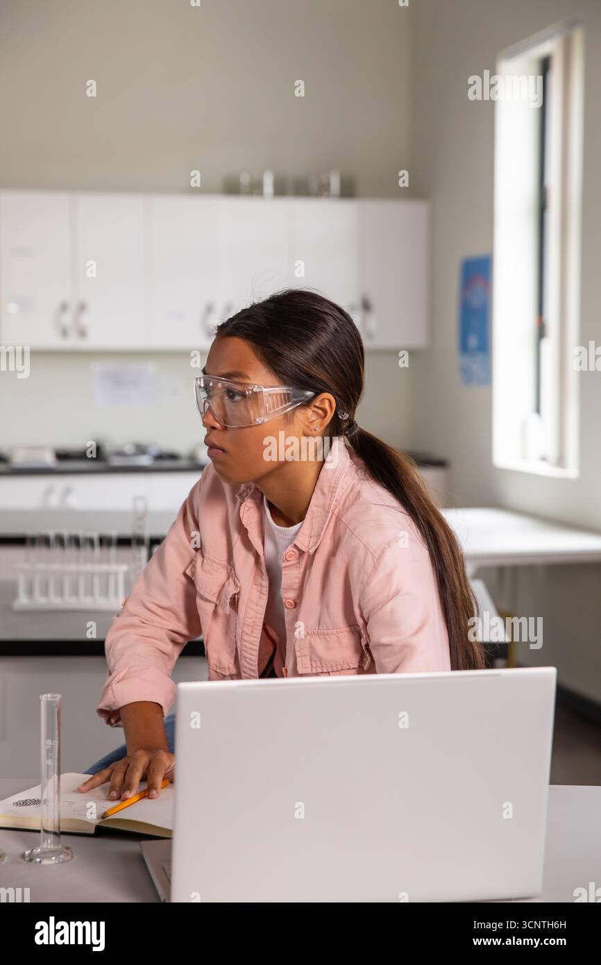 Chinese female student wearing goggles writing in notebook at lab bench ...