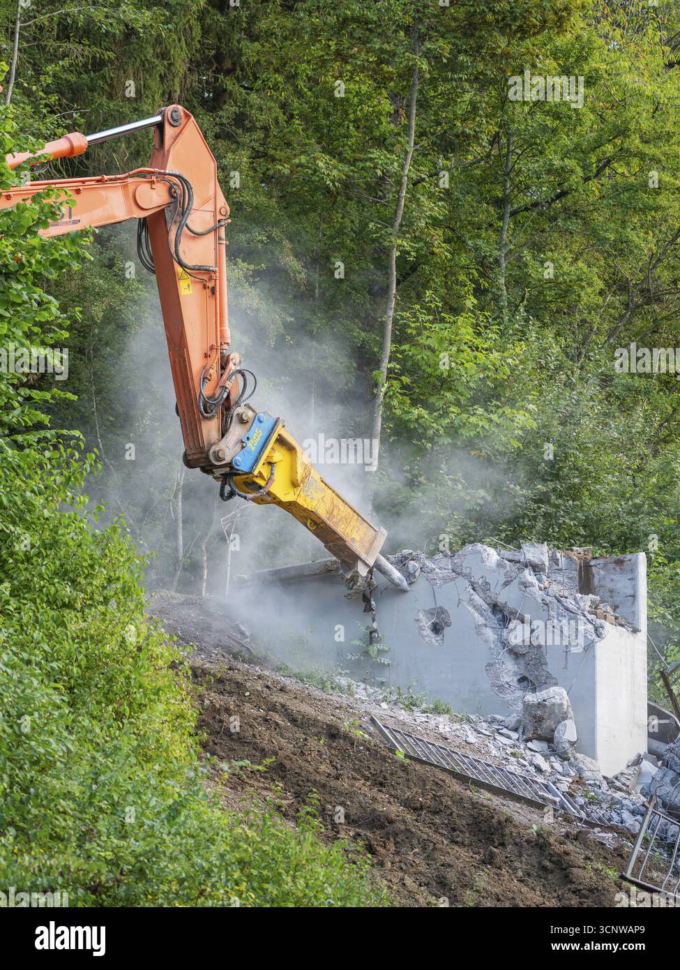 An excavator breaks a concrete wall in a green environment, dust rises ...