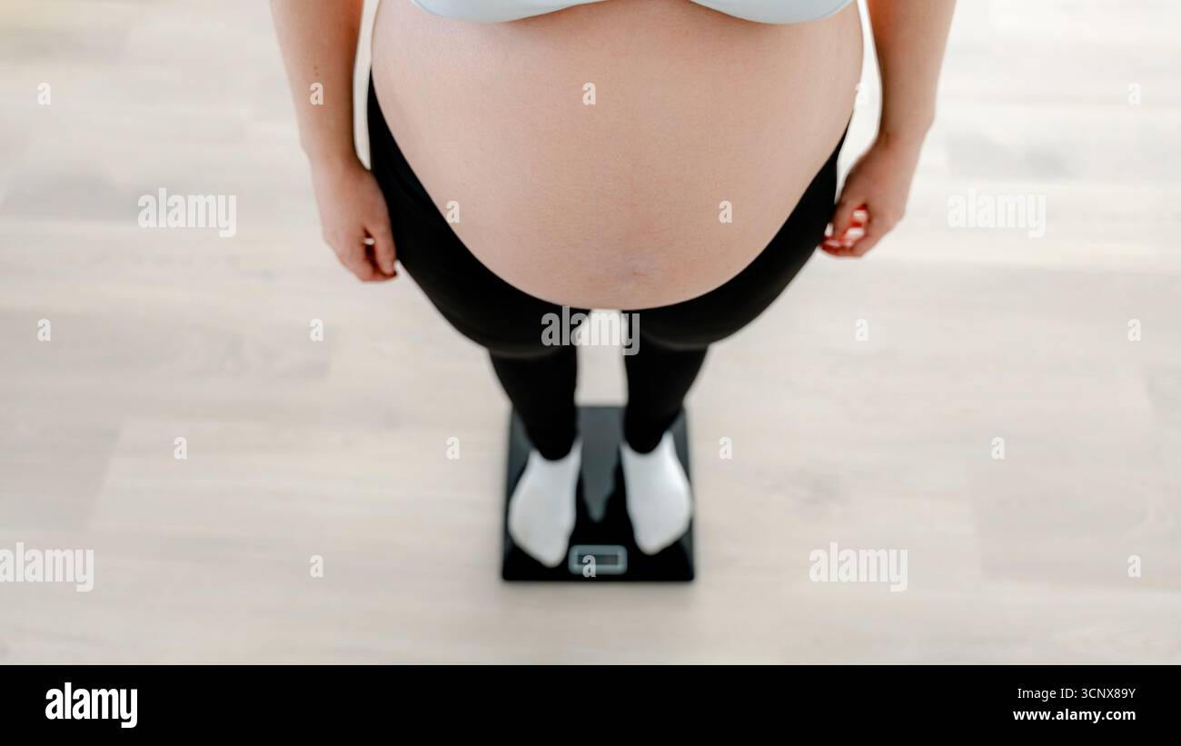 A pregnant woman stands on a digital scale, monitoring her weight ...