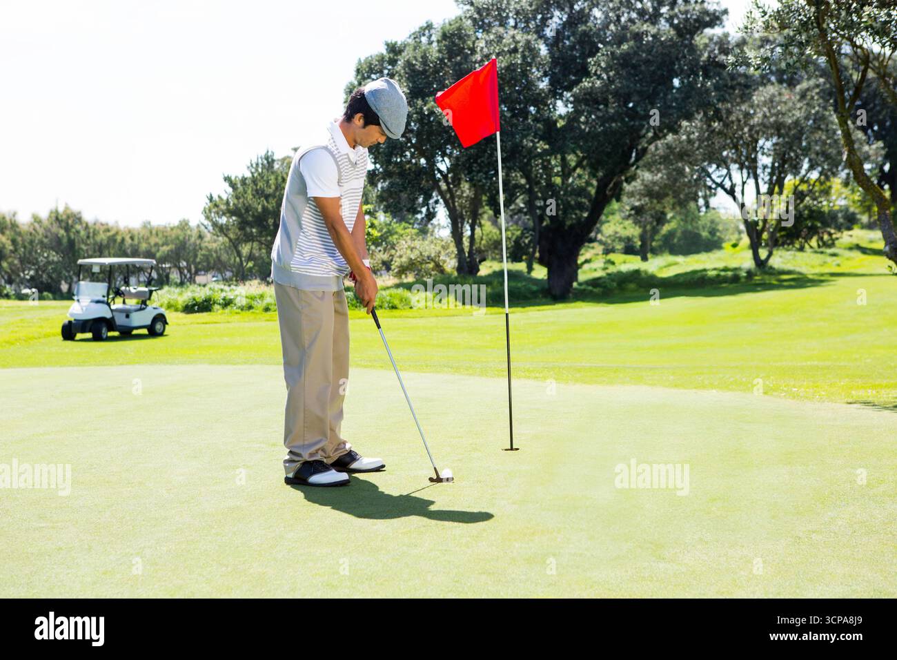 Asian man aligning putter with golf ball on green by red flag with golf ...