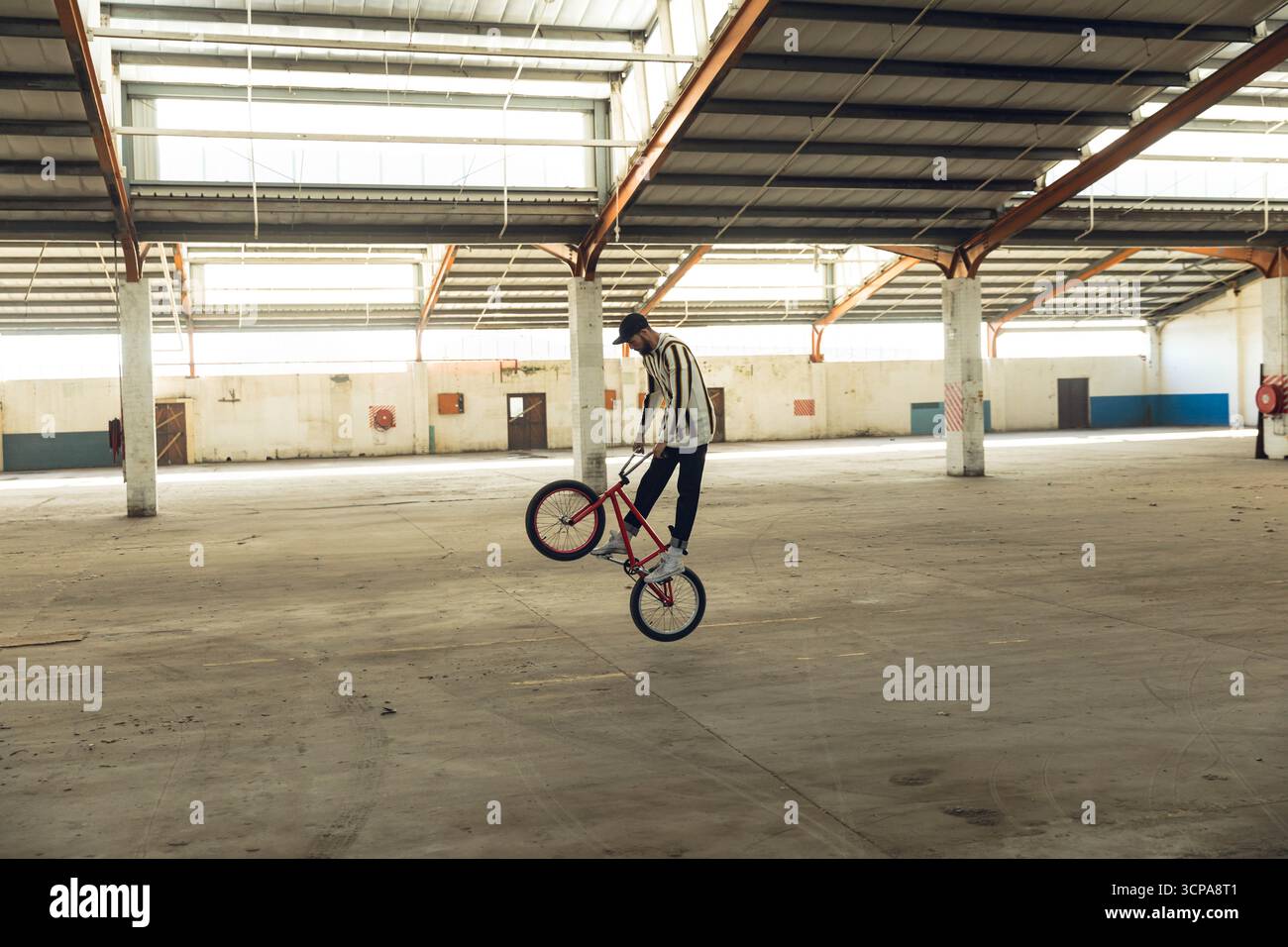 Male BMX rider balancing on rear wheel beneath exposed trusses inside ...