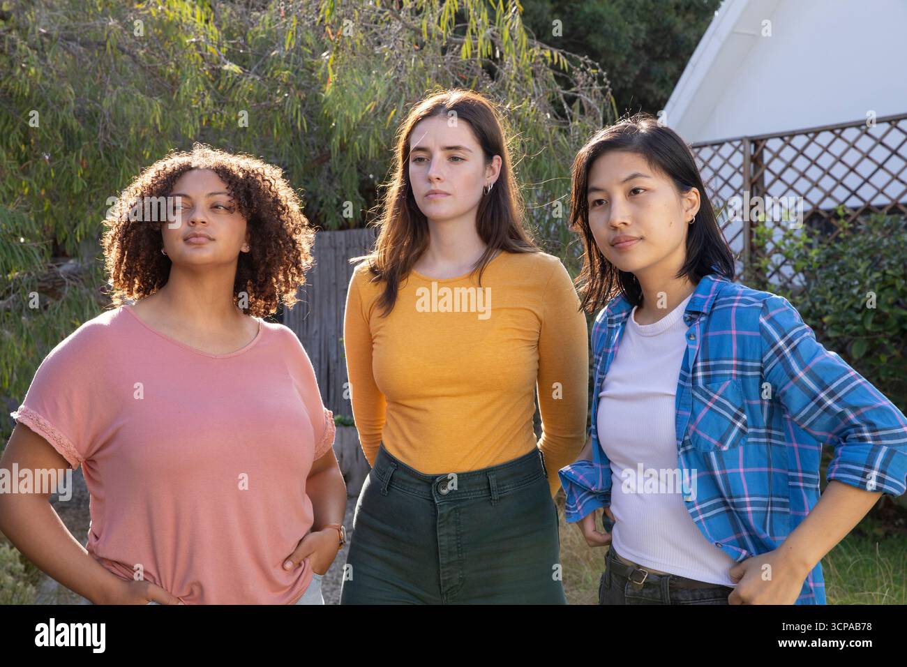 Diverse female friends standing in backyard amid garden shrubs, lattice ...