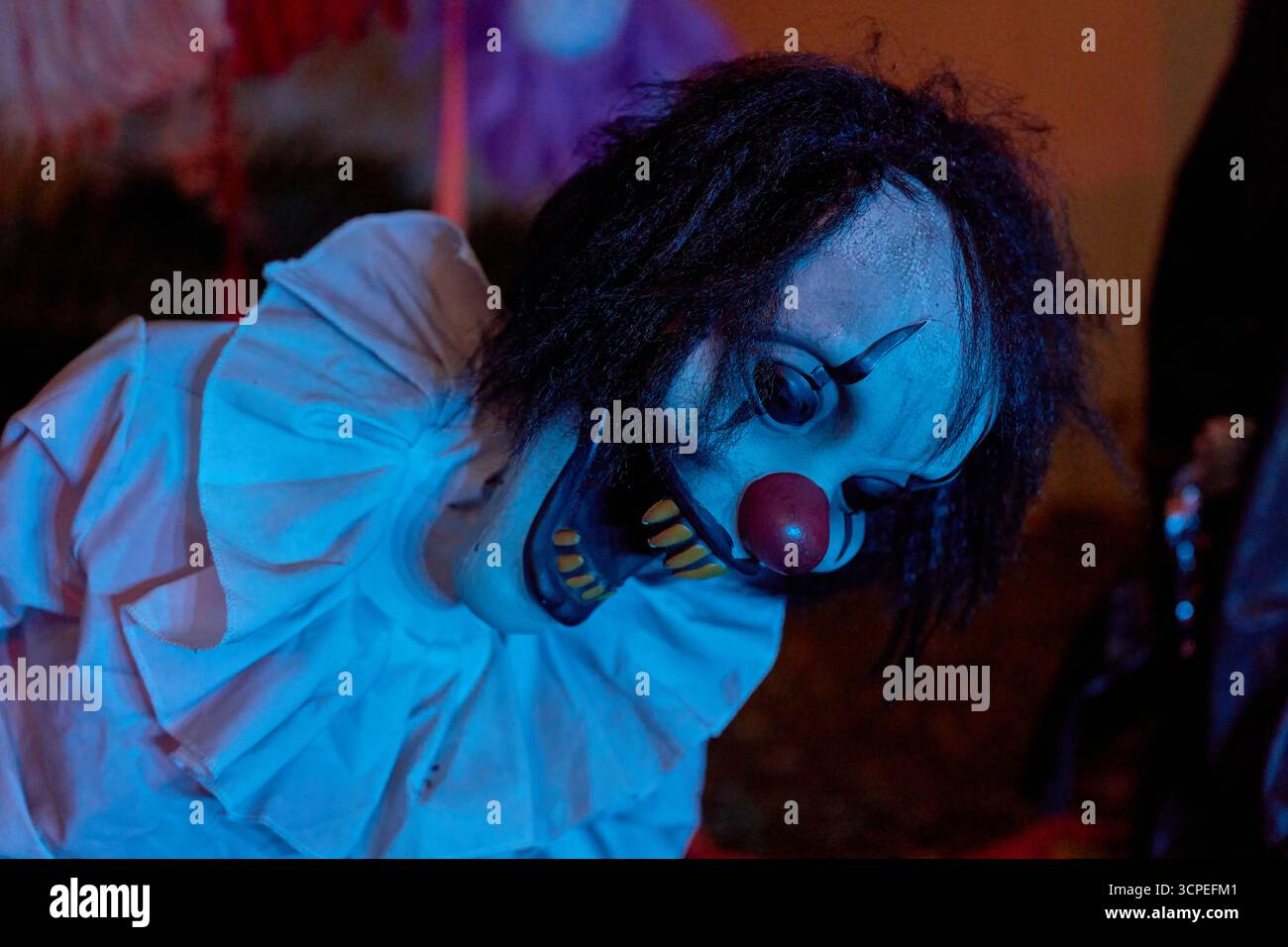 A scary clown in a white ruffled costume leans forward under blue light ...