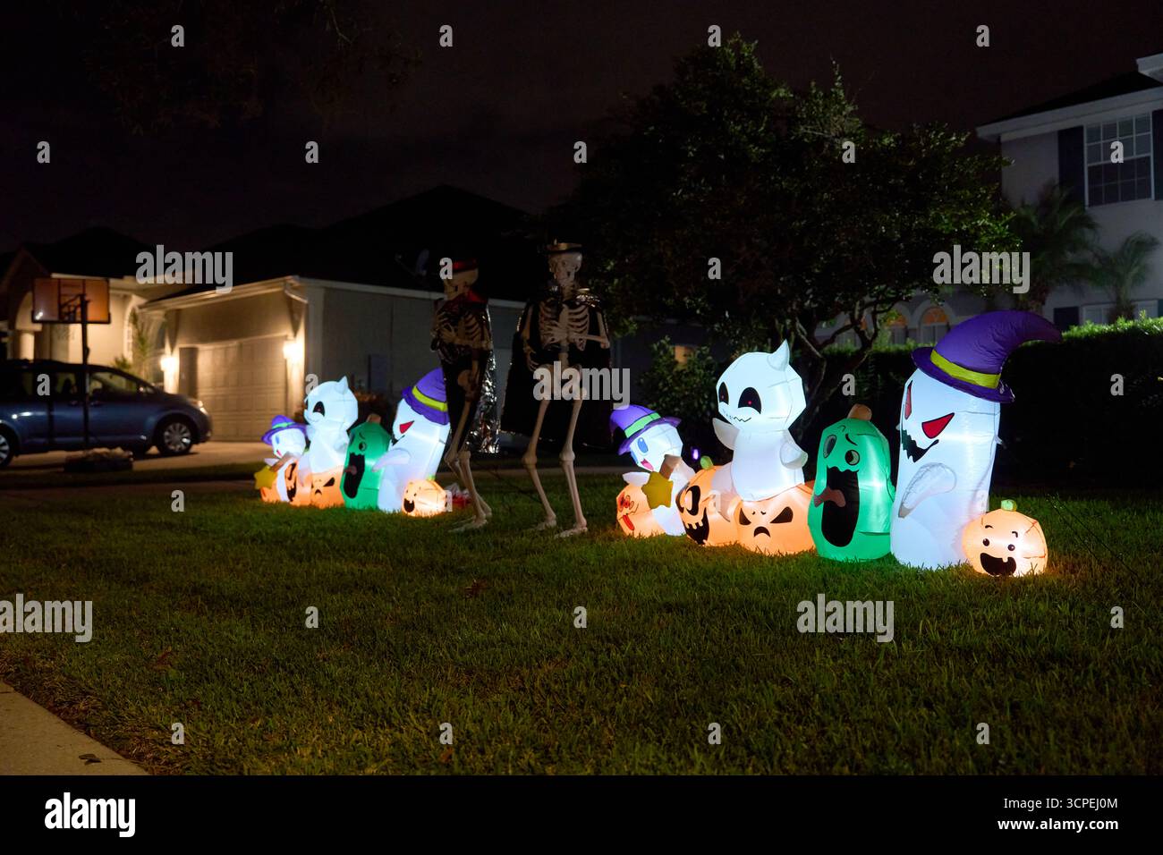 A row of glowing inflatable ghosts and pumpkins lines a suburban lawn ...