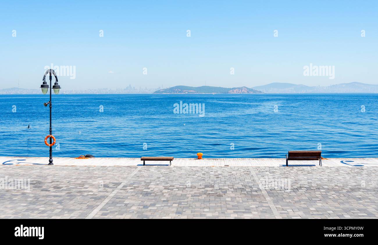 Two wooden benches sit along a paved promenade by the water. The calm ...