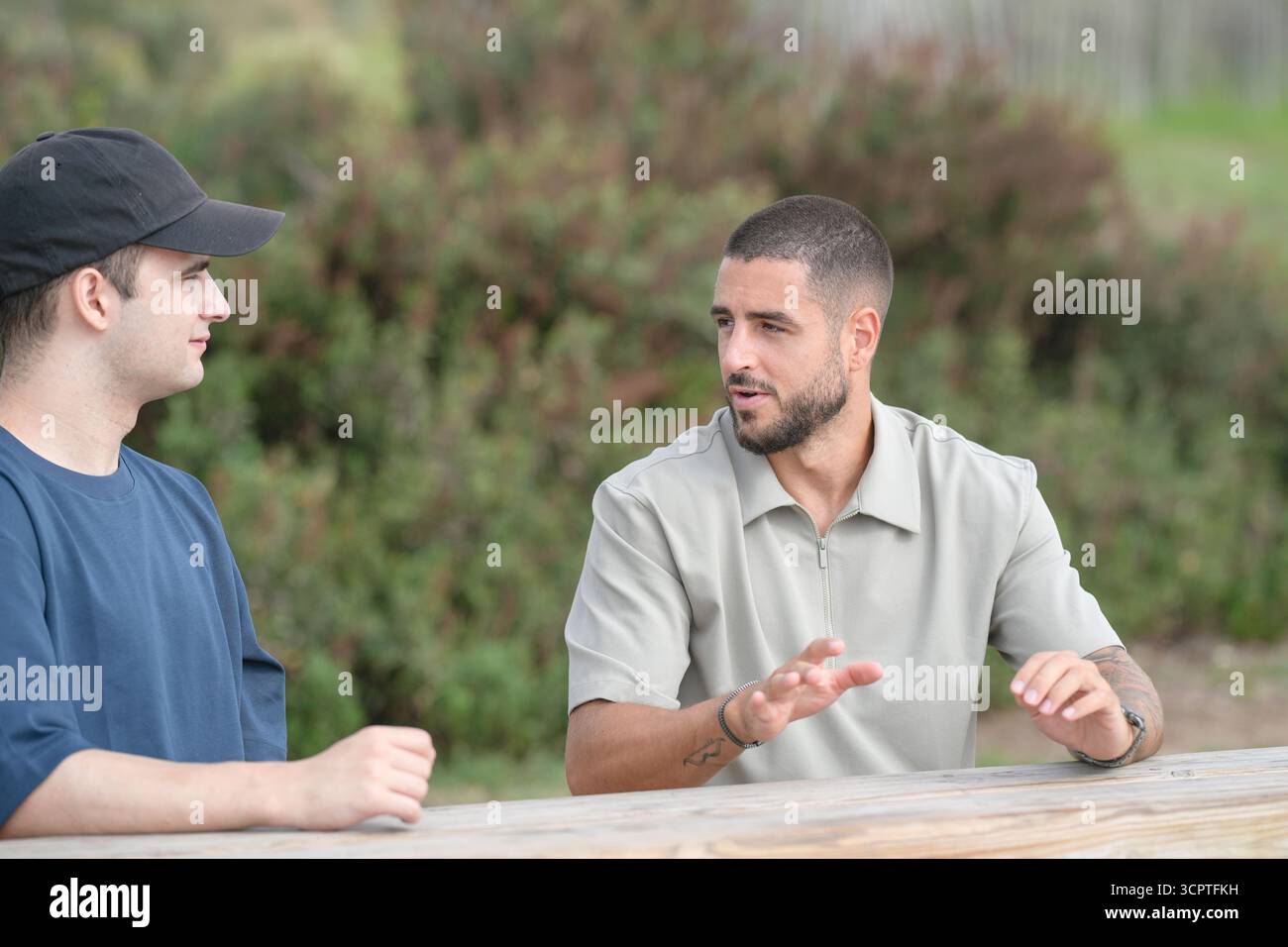 Two men are sitting at a picnic table, one wearing a black hat. They ...