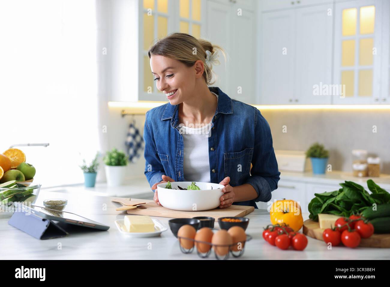 Smiling woman looking at recipe on tablet while cooking at table in ...