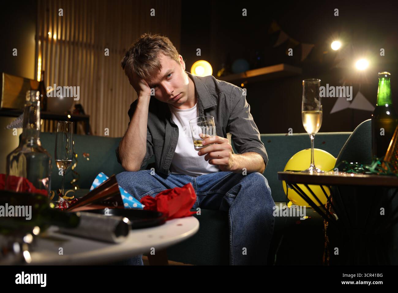 Man with glass of alcohol suffering from hangover on sofa in messy room ...