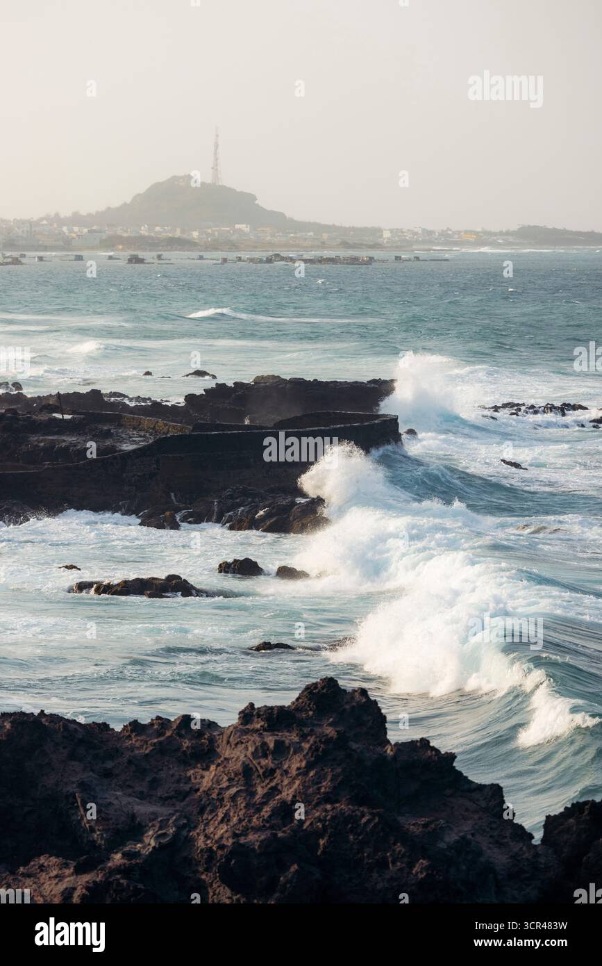 Rugged rocky coastline with crashing waves under a hazy sky and distant ...