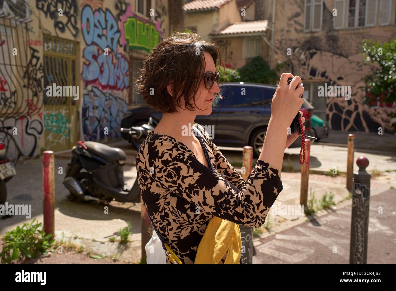 Woman with sunglasses takes a photo in a graffiti-covered urban street ...