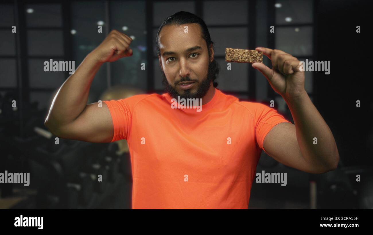 Man flexes bicep while holding a protein snack bar by his face in a gym ...