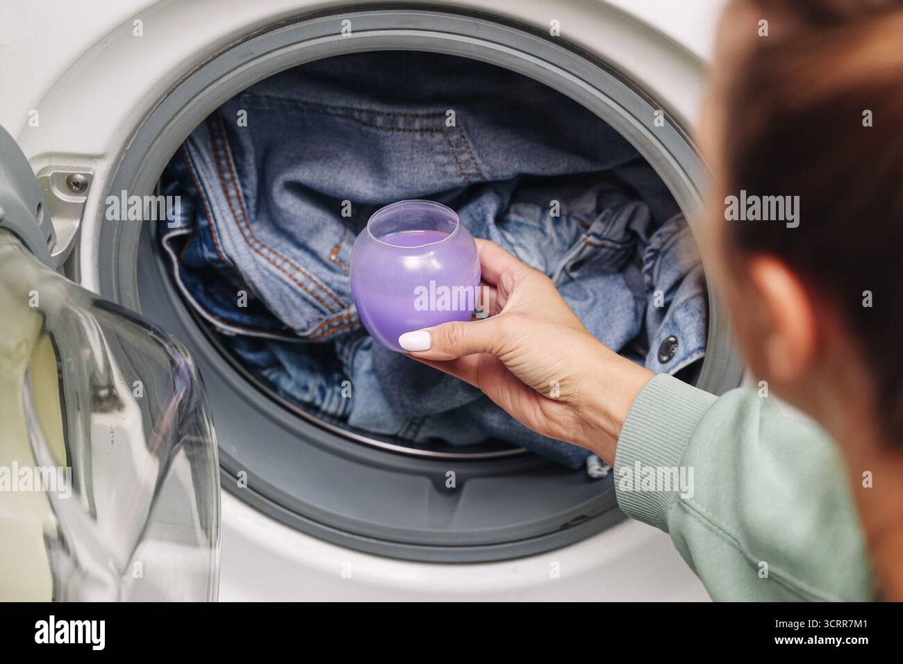 Woman loading denim jeans into washing machine with purple detergent ...