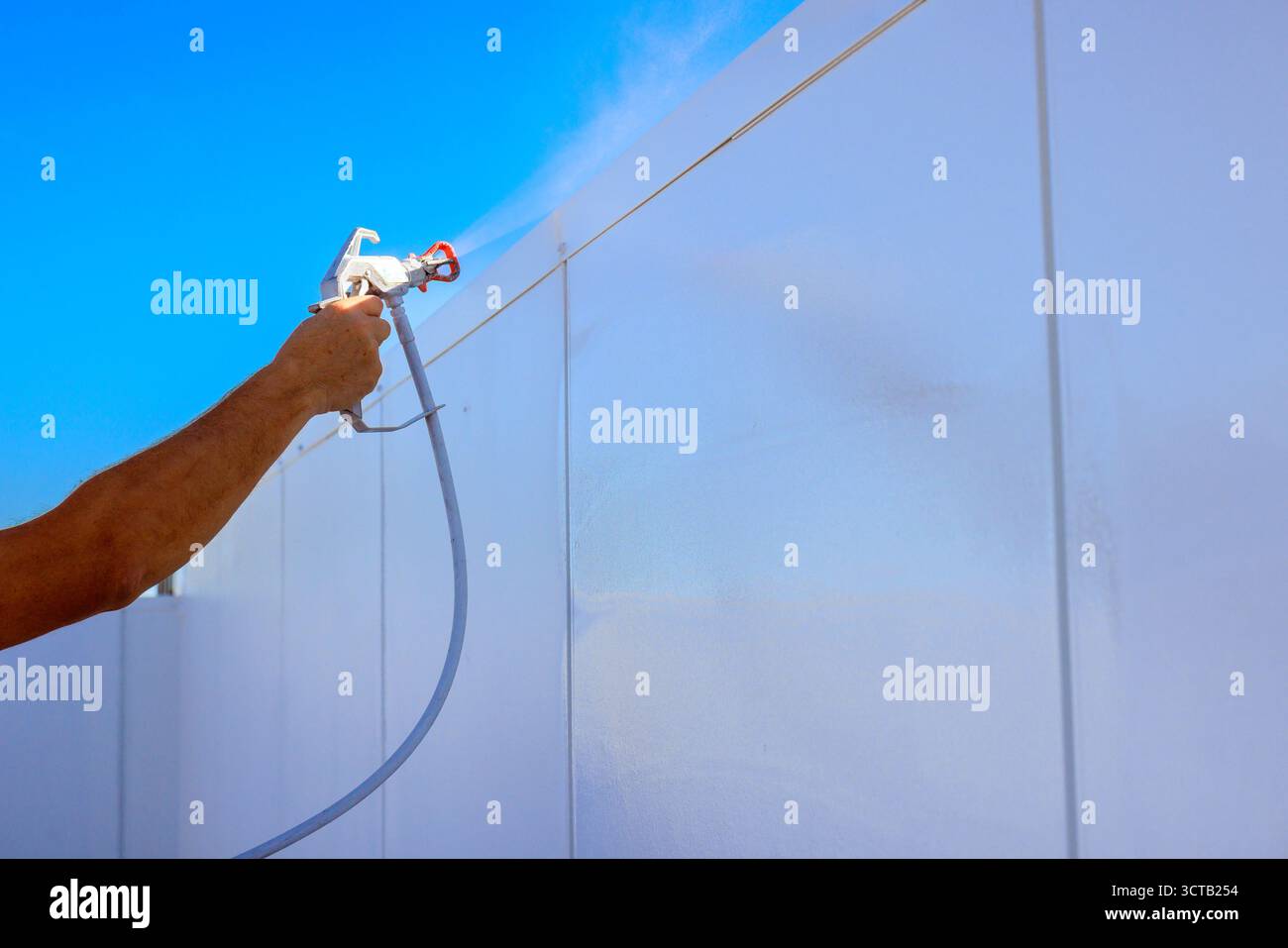 Worker is painting tall white wall with paint spray gun while workday ...