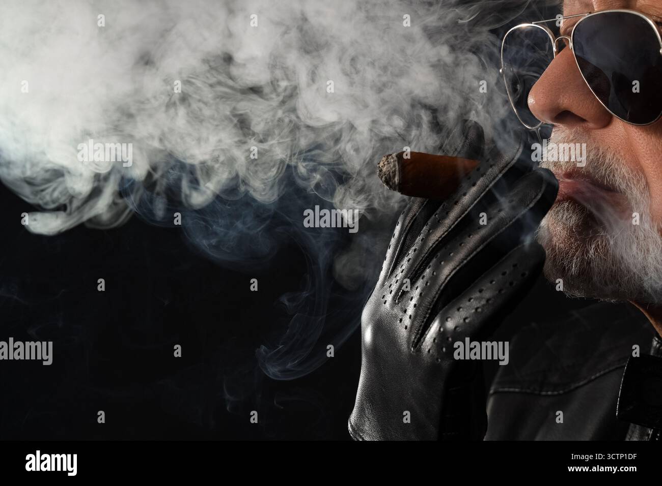 Stylish old man in leather jacket smoking cigar on black background ...
