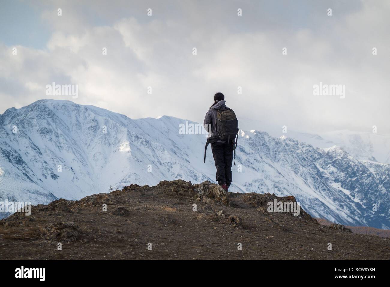 Hiker with backpack gazes across rugged cliff toward vast snow capped ...