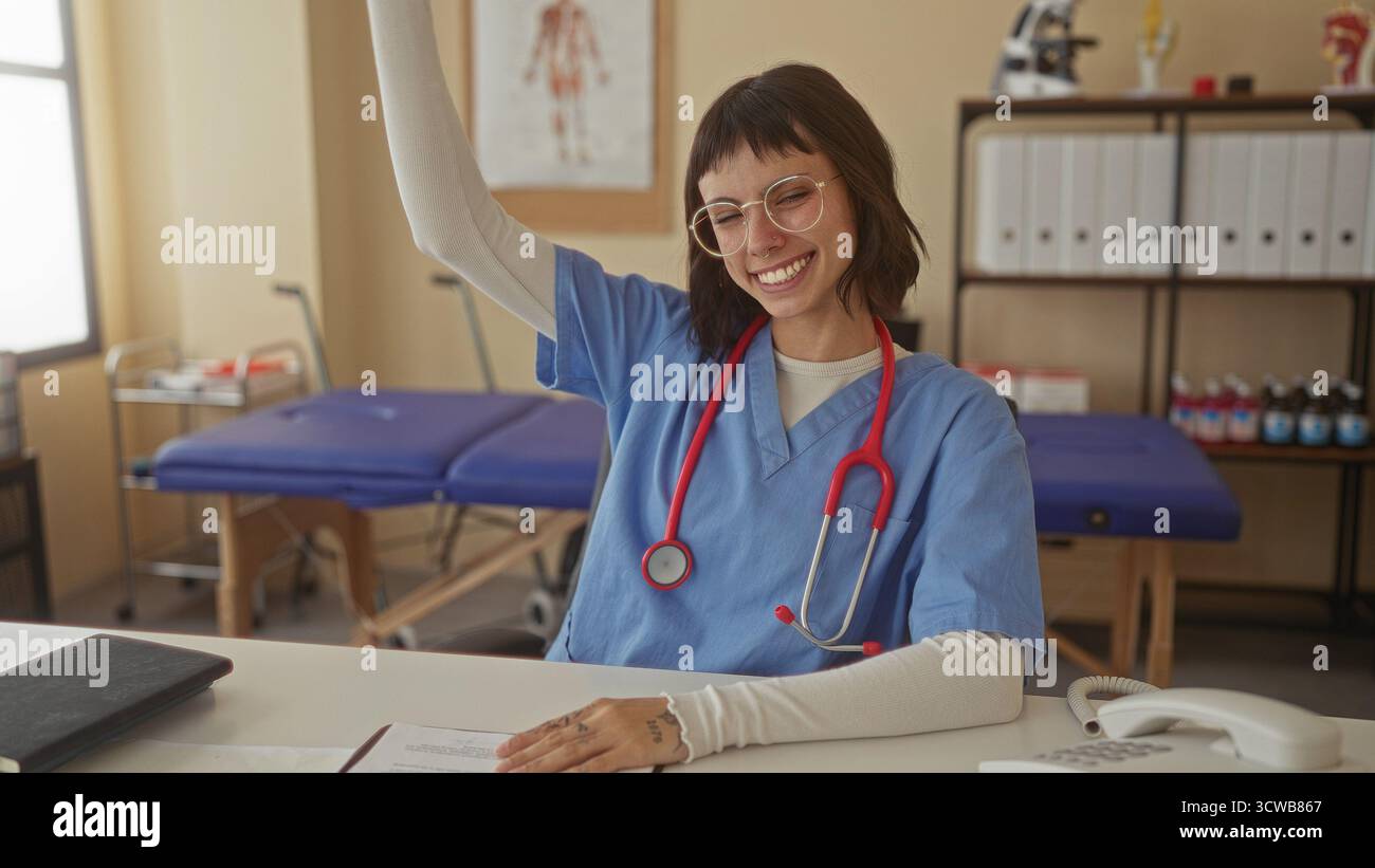 Woman nurse wearing blue scrubs and stethoscope raises arm in building ...