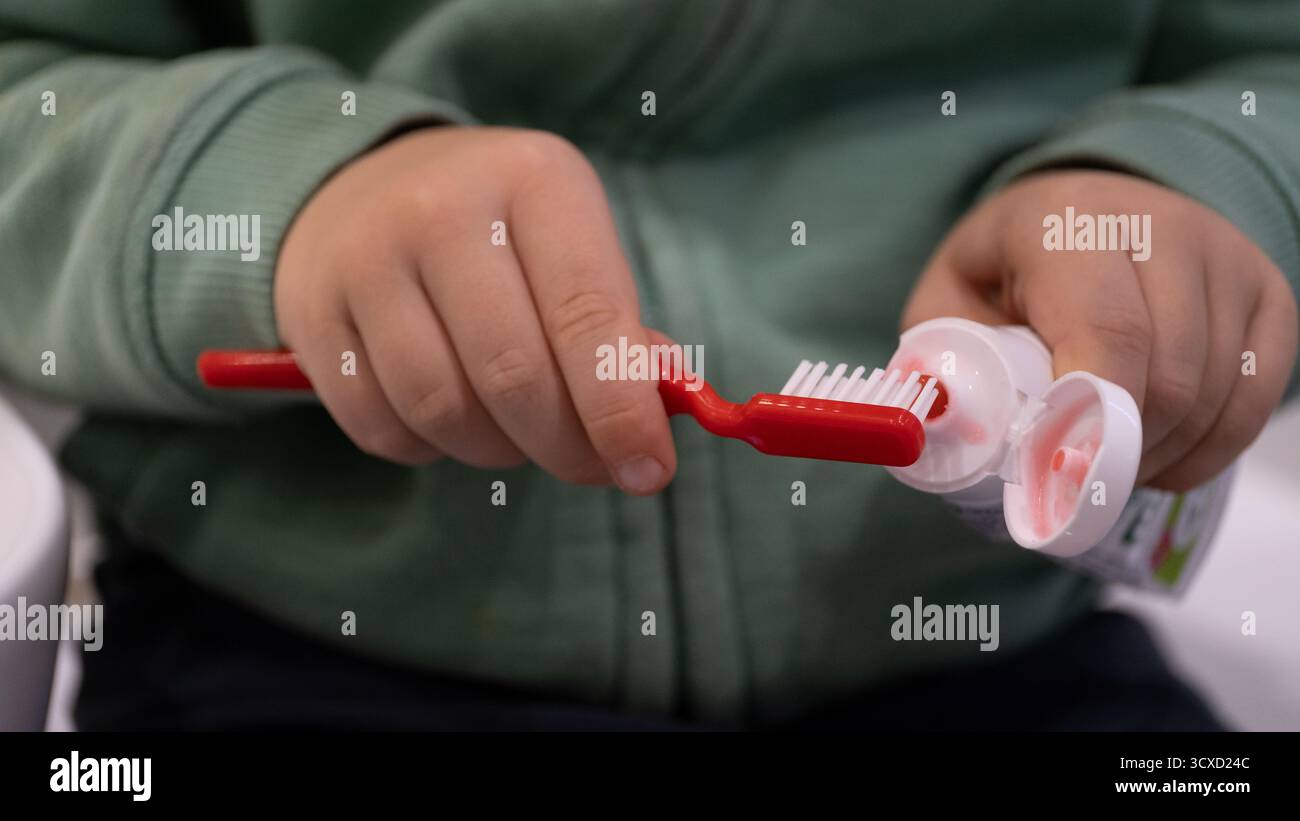 Child squeezing toothpaste onto a red toothbrush during morning hygiene ...