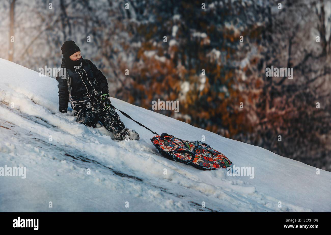A boy dressed in warm winter clothes sits on a snowy hill beside a ...