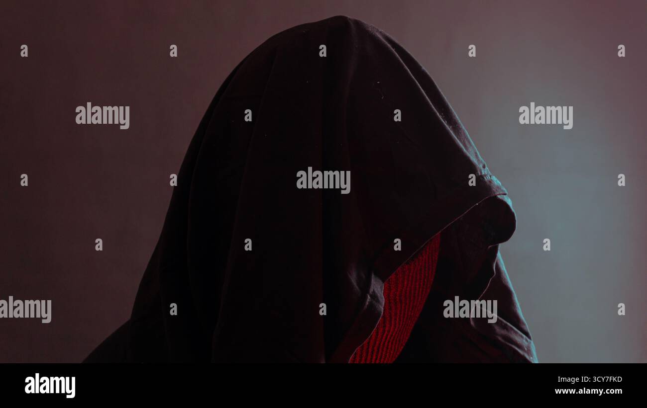 Hooded and masked figure under red ambient light against dark backdrop ...