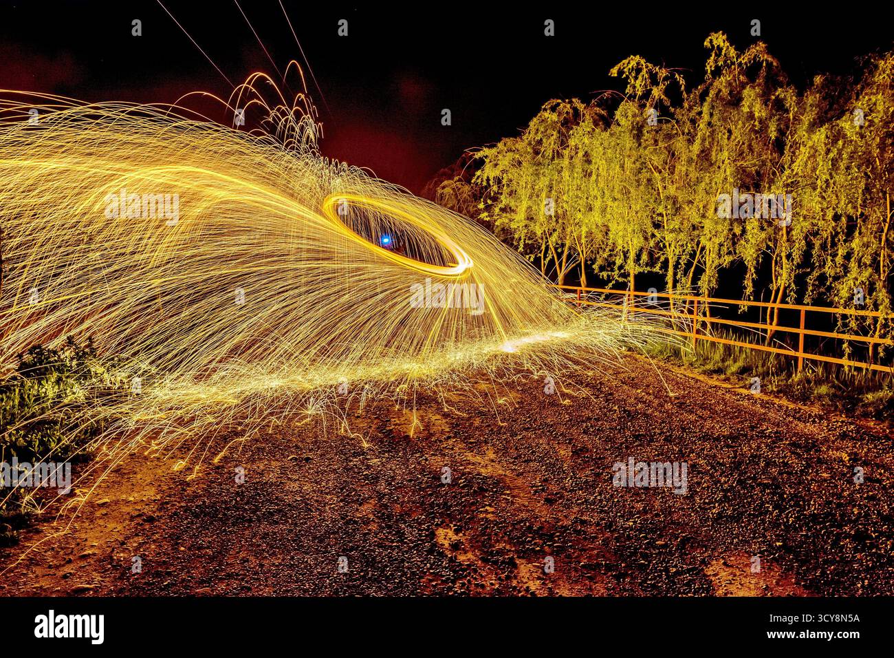 Steel wool spinning creates vortex of golden sparks. Trees and railing ...