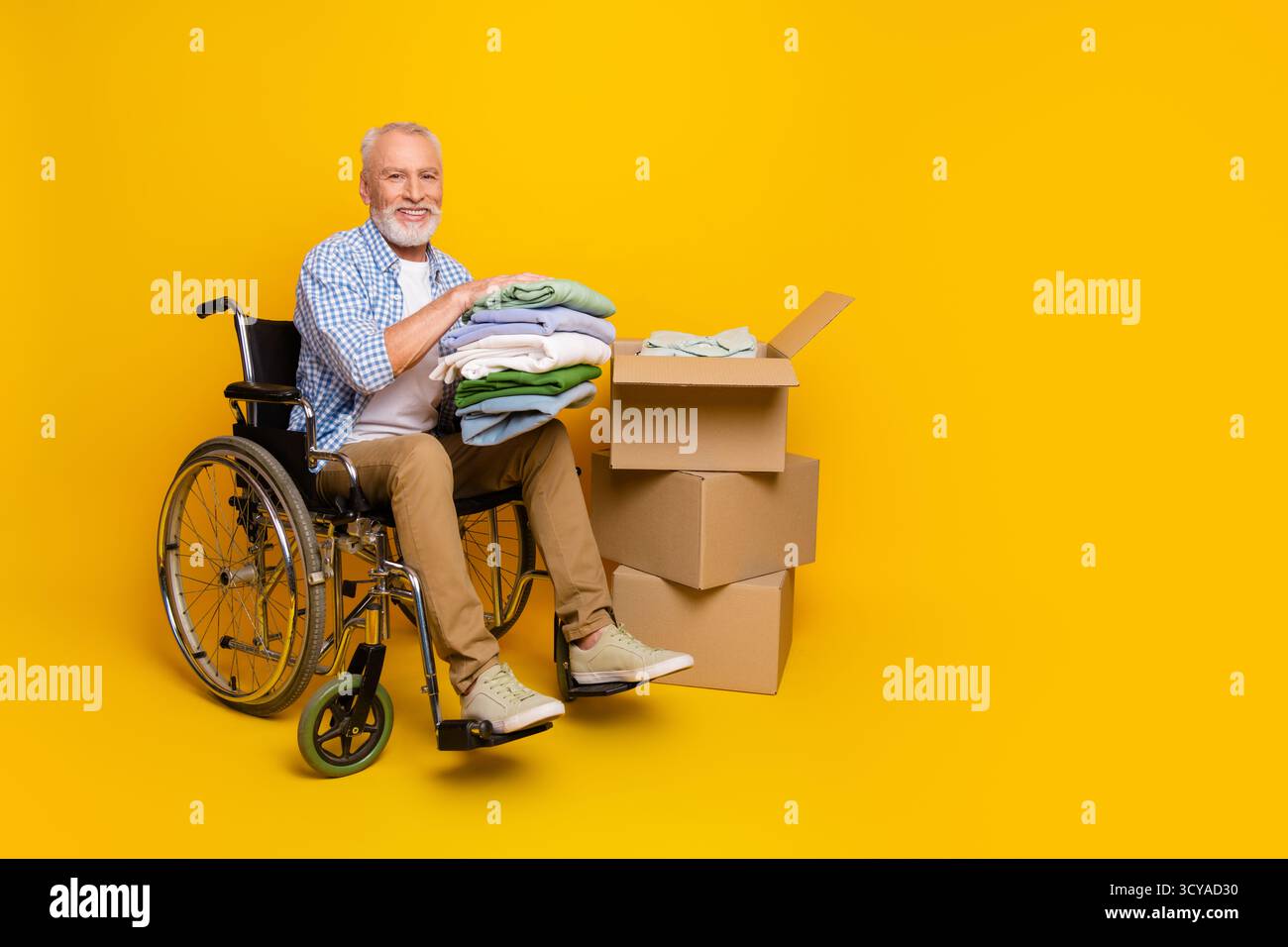 Funny grandpa in a wheelchair packs colorful clothes beside cardboard ...