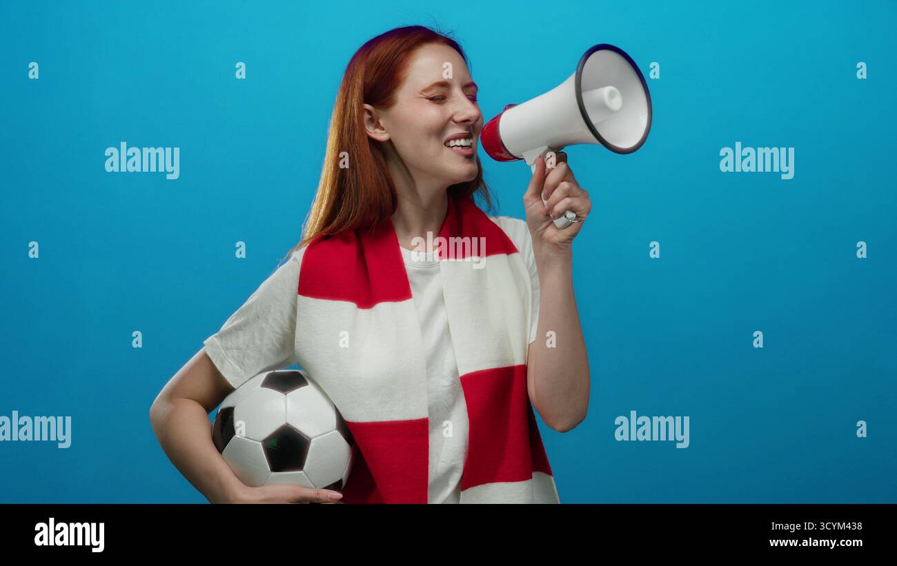 Woman cheering with megaphone while holding soccer ball against blue ...