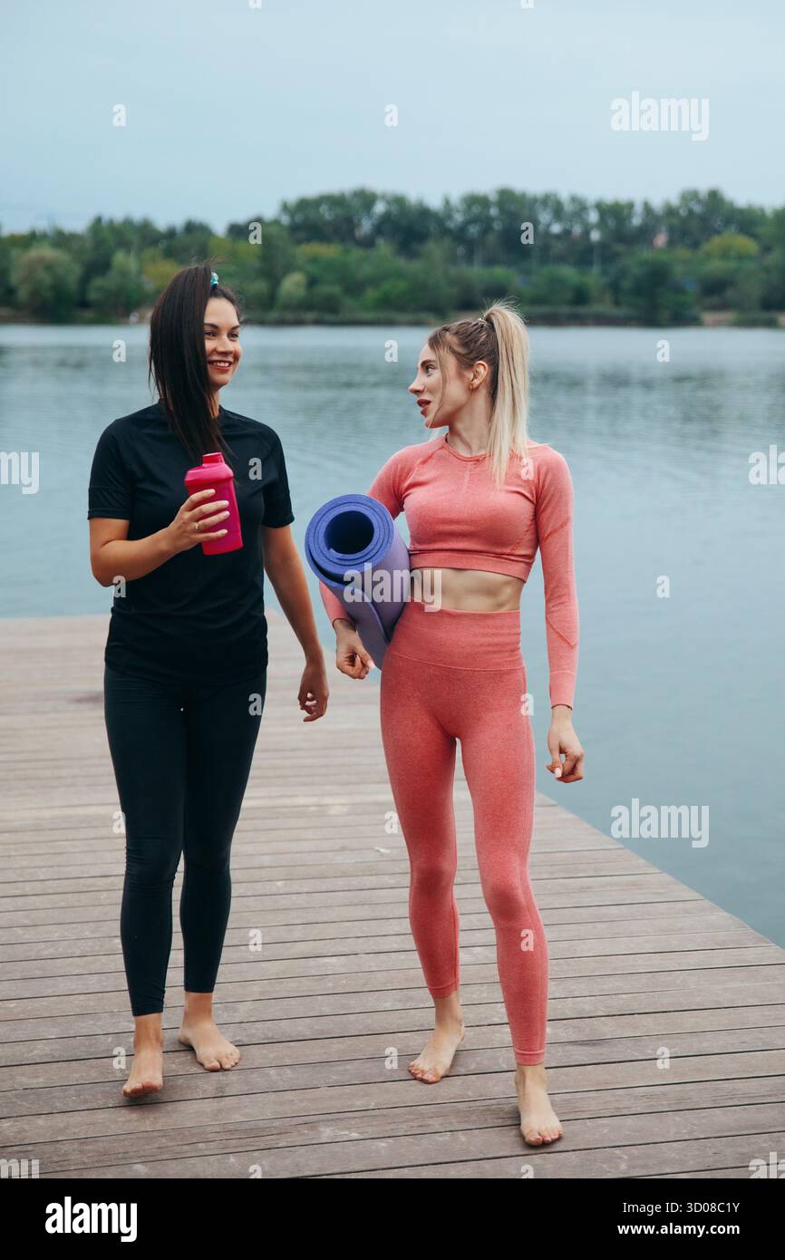 Two women walk together by a calm lake, holding yoga props and ...