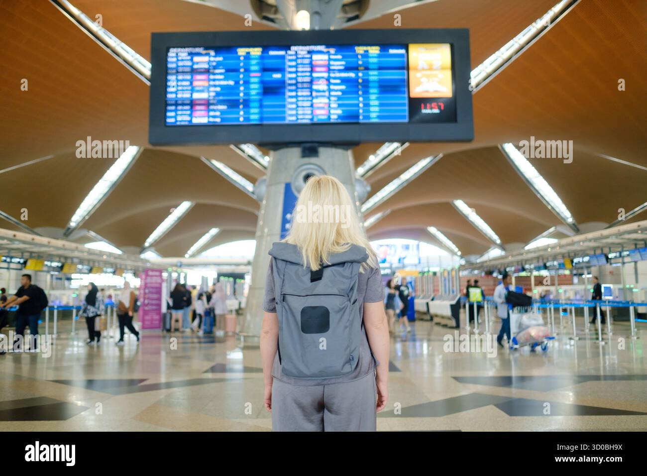 A traveler stands in an airport terminal, facing a digital flight ...
