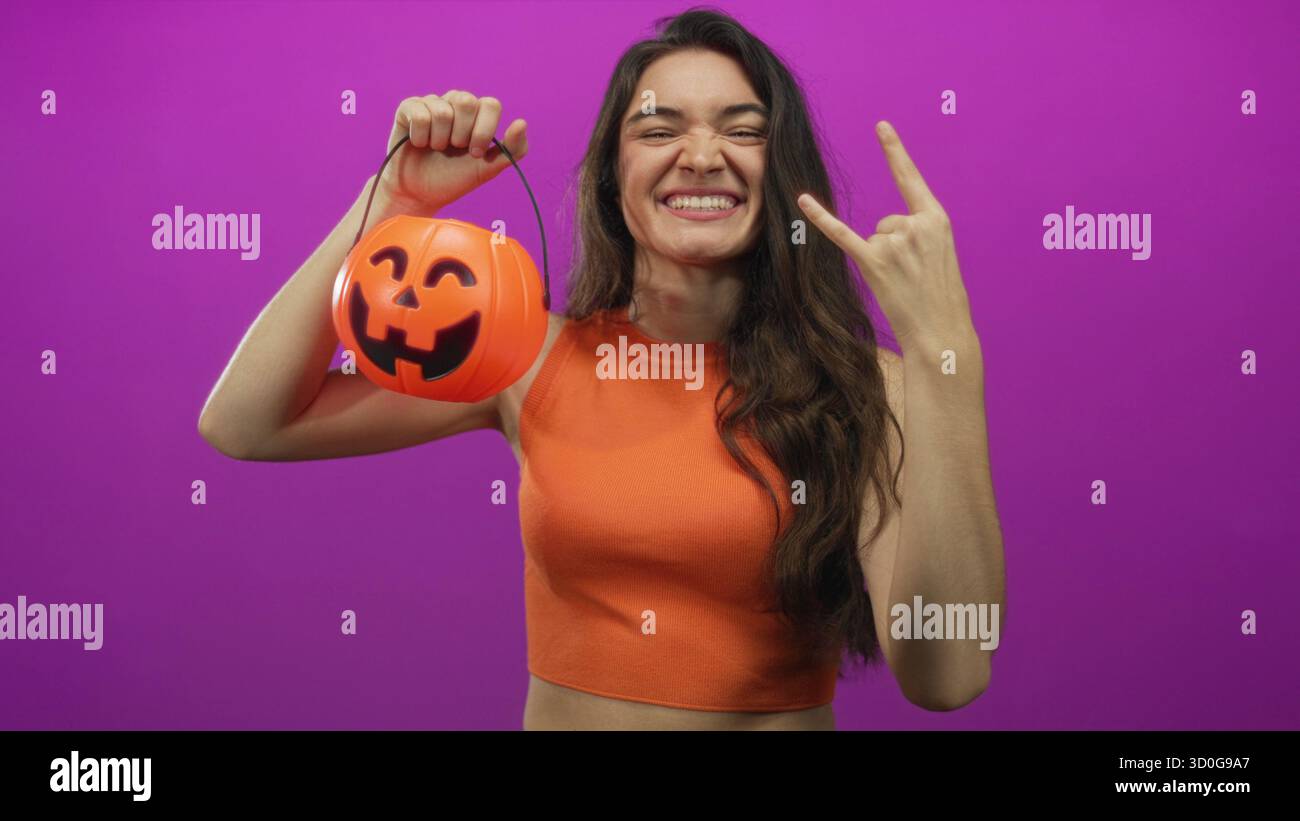 Young hispanic woman holding a pumpkin bucket and making rock horns ...