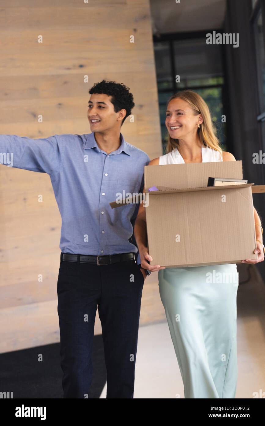 Colleagues smiling while carrying box and pointing in modern office ...