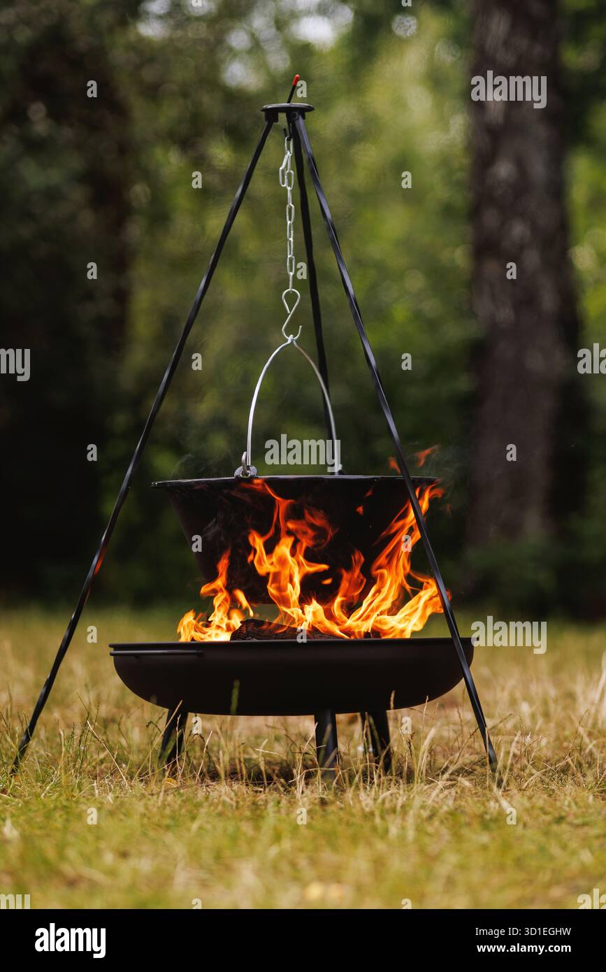 Metal pot hanging on tripod over open campfire in grassy field with ...
